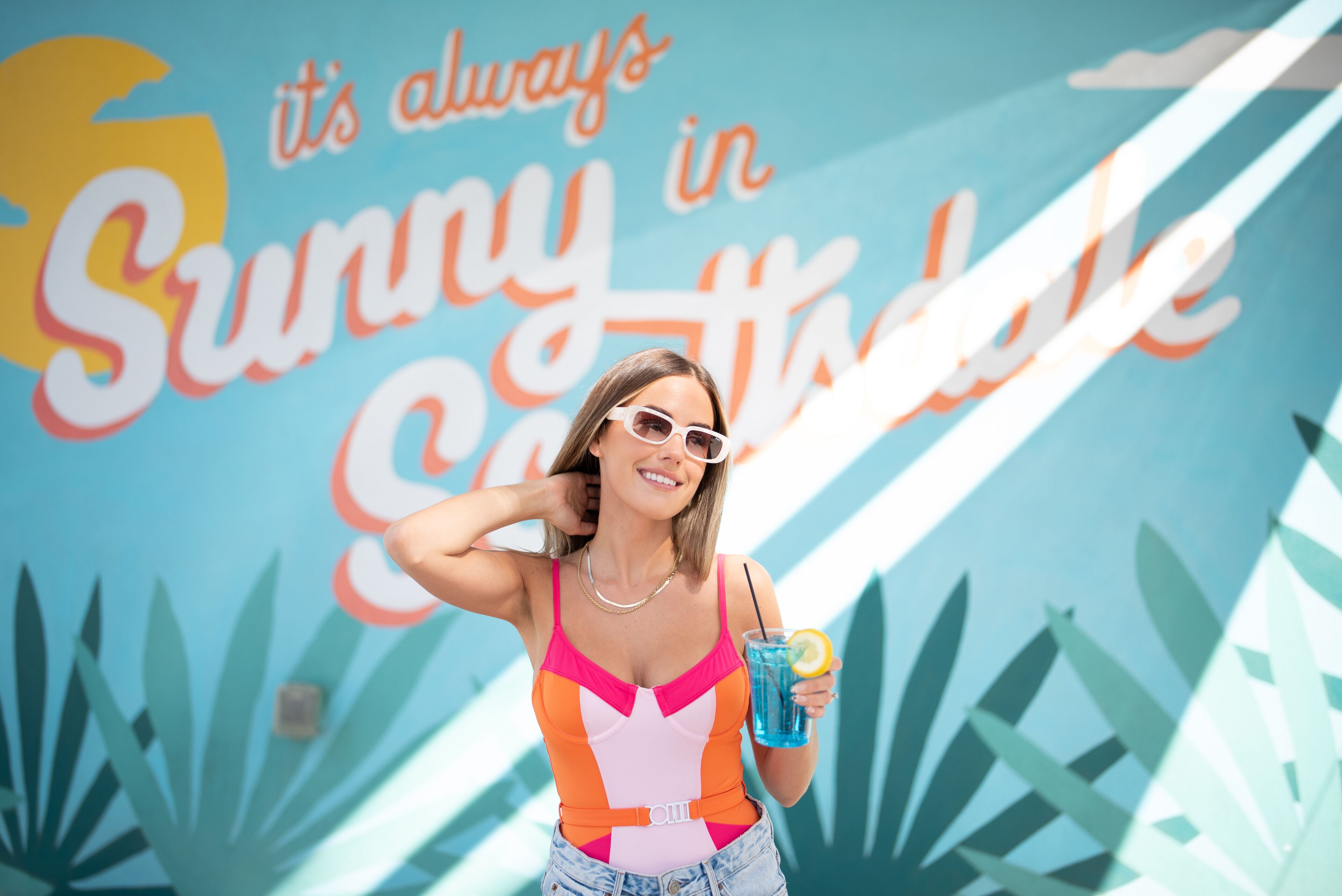 Uma pessoa de fato de banho colorido segura uma bebida com uma fatia de limão, em frente a uma parede com o texto "it's always sunny in Sunnydale".