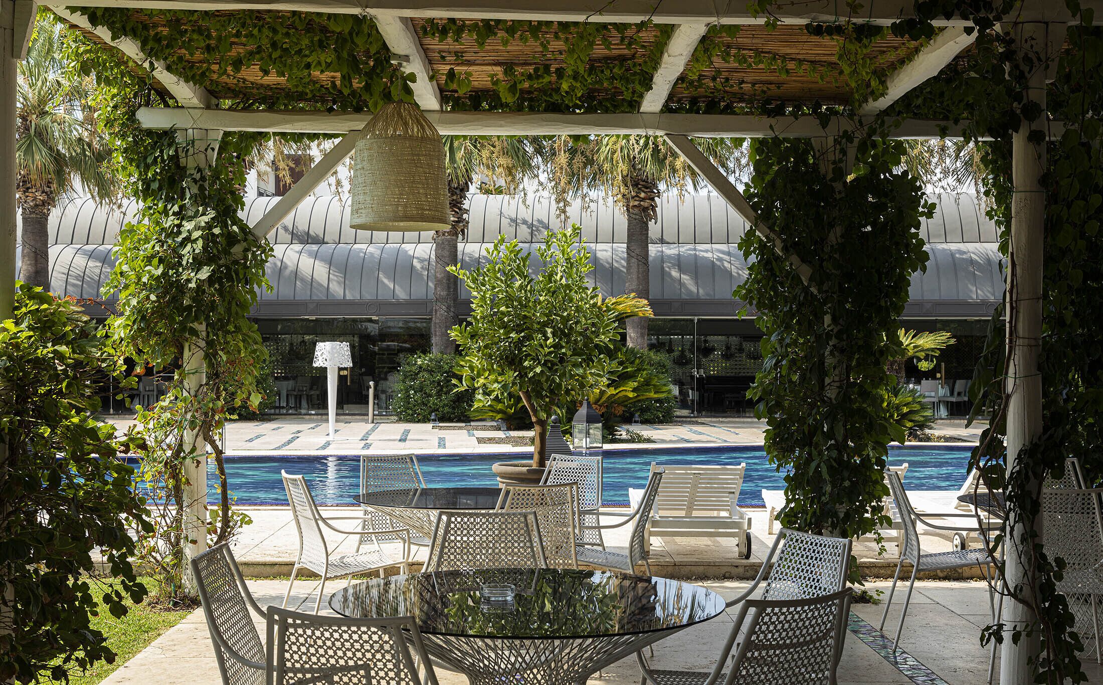Uma área de estar coberta ao ar livre com vista para a piscina, cadeiras de metal, mesa de vidro e uma pérgula com plantas trepadeiras.