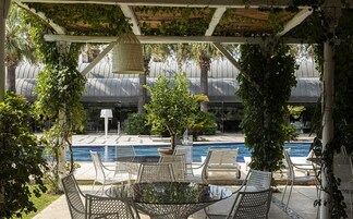 Uma área de estar coberta ao ar livre com vista para a piscina, cadeiras de metal, mesa de vidro e uma pérgula com plantas trepadeiras.