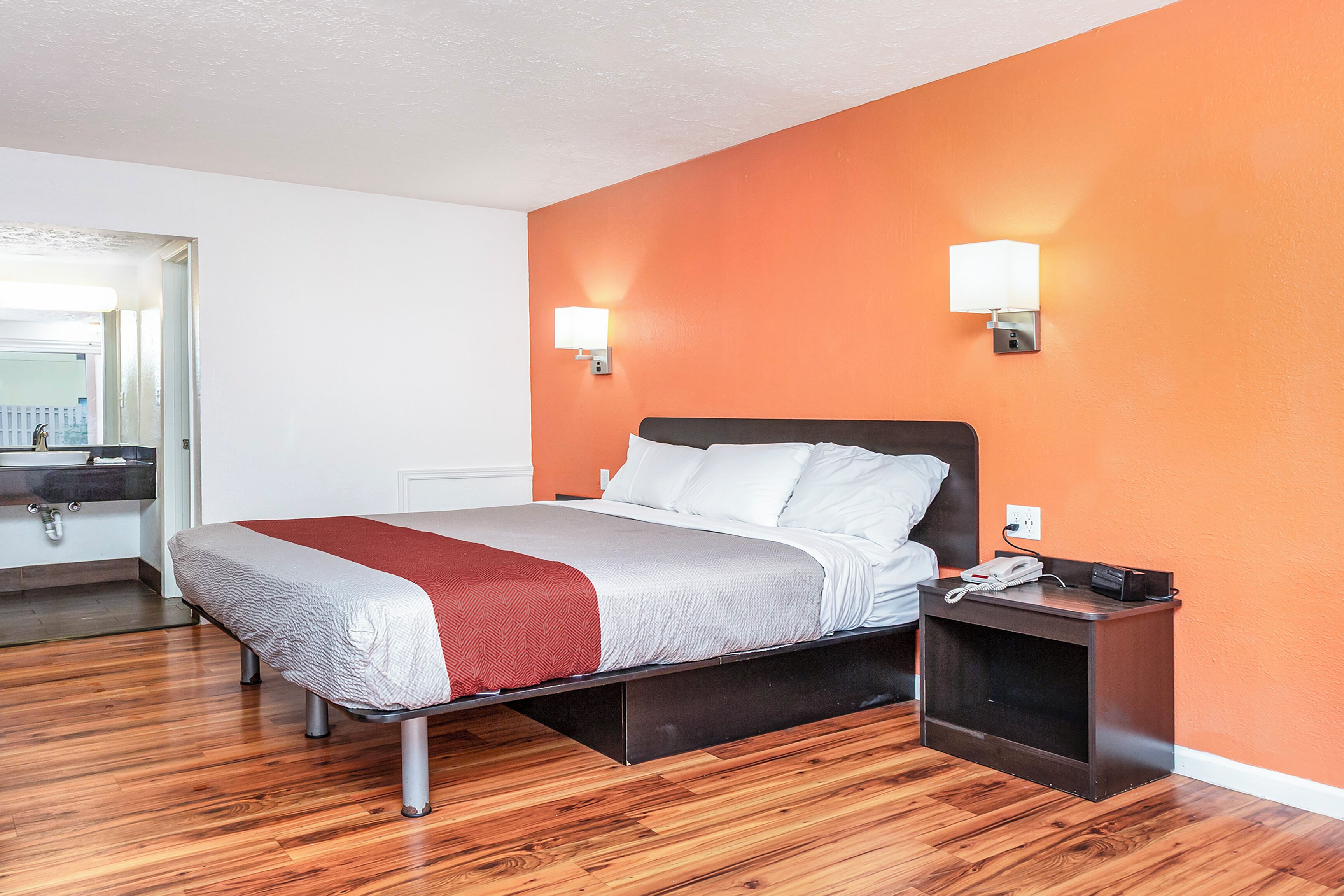 Quarto de hotel moderno com piso de madeira, cama com colcha cinza e vermelha, duas luminárias fixadas na parede e mesa de cabeceira com telefone e carregador.