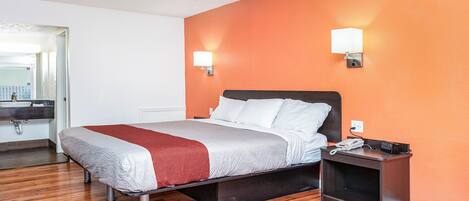 Quarto de hotel moderno com piso de madeira, cama com colcha cinza e vermelha, duas luminárias fixadas na parede e mesa de cabeceira com telefone e carregador.
