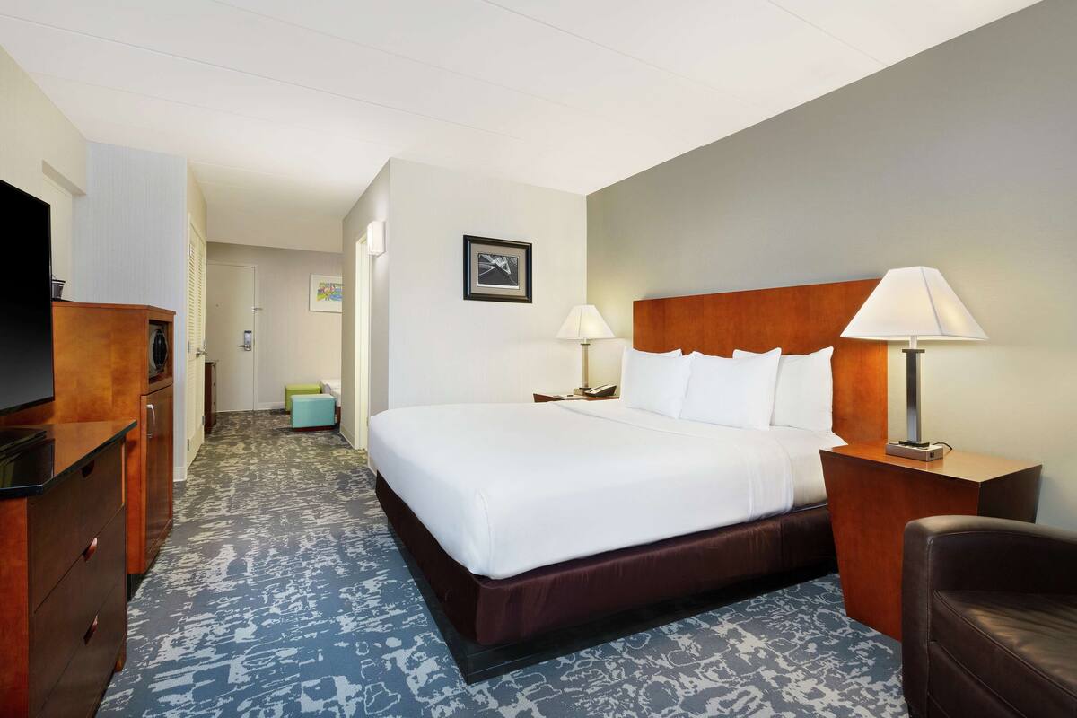 Quarto de hotel com uma cama grande, duas luminárias de cabeceira, uma cabeceira de madeira, uma televisão e uma escrivaninha com gavetas.