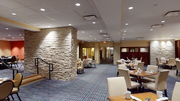 Uma área de jantar de hotel com várias mesas e cadeiras, uma parede de destaque em pedra e piso coberto de carpete.