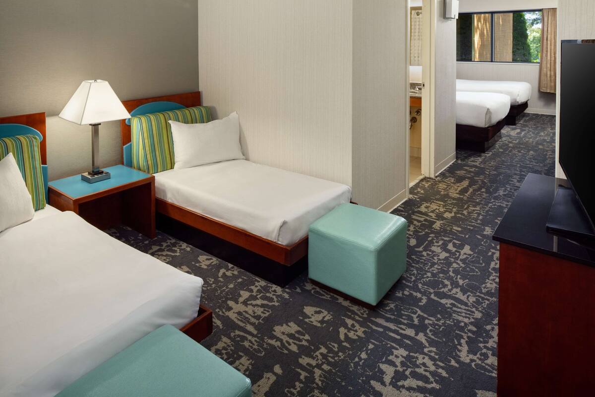 Quarto de hotel com duas camas de solteiro, uma escrivaninha com luminária, televisão e banheiro visível por uma porta aberta.