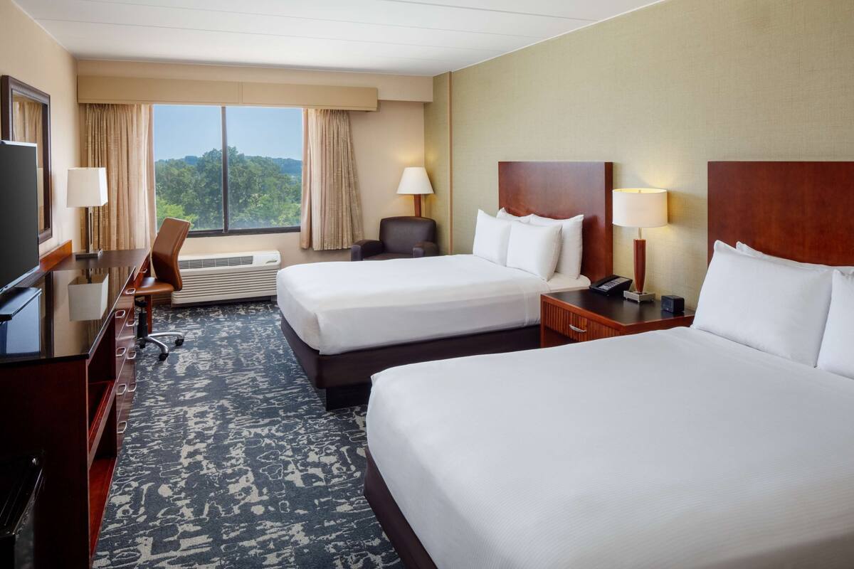 Quarto de hotel com duas camas, uma mesa, uma cadeira, uma TV e janela com vista para o exterior.