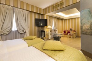 Quarto de hotel com uma cama grande, televisão, área de estar com sofá e poltronas, e um quadro na parede.