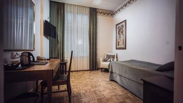 Quarto de hotel com piso de madeira em padrão herringbone, uma cama, uma escrivaninha com cadeira, uma televisão na parede e uma janela com cortinas.