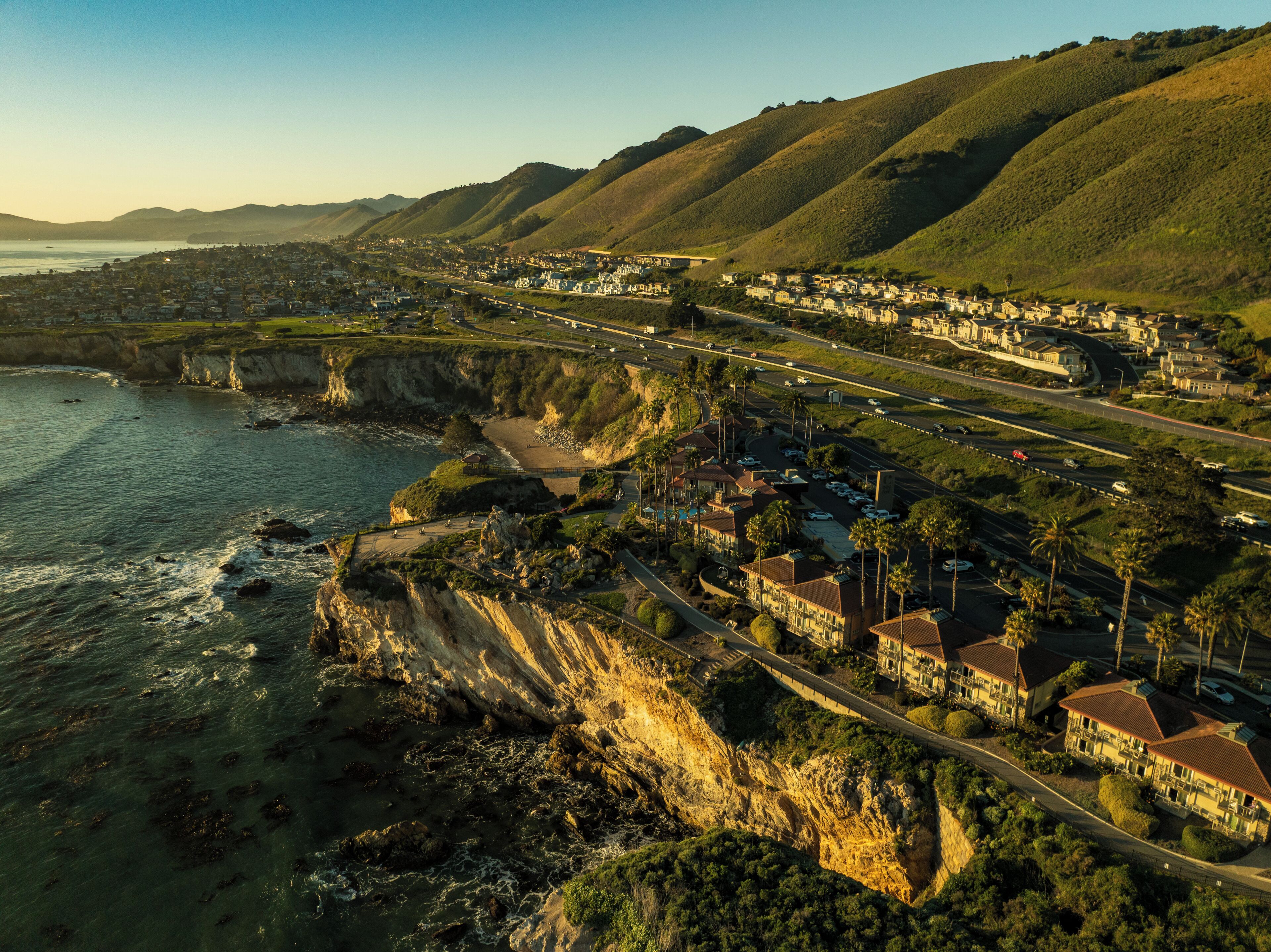 Uma vila costeira com um penhasco imponente sobre o oceano, uma estrada principal atravessando o centro e edifícios residenciais com varandas visíveis.