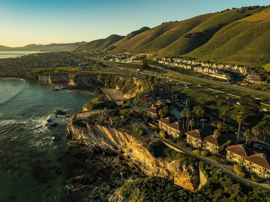 Uma vila costeira com um penhasco imponente sobre o oceano, uma estrada principal atravessando o centro e edifícios residenciais com varandas visíveis.