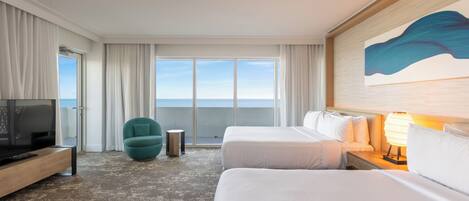 Quarto de hotel com duas camas, televisão, uma cadeira verde-água e uma janela grande com vista para o oceano.