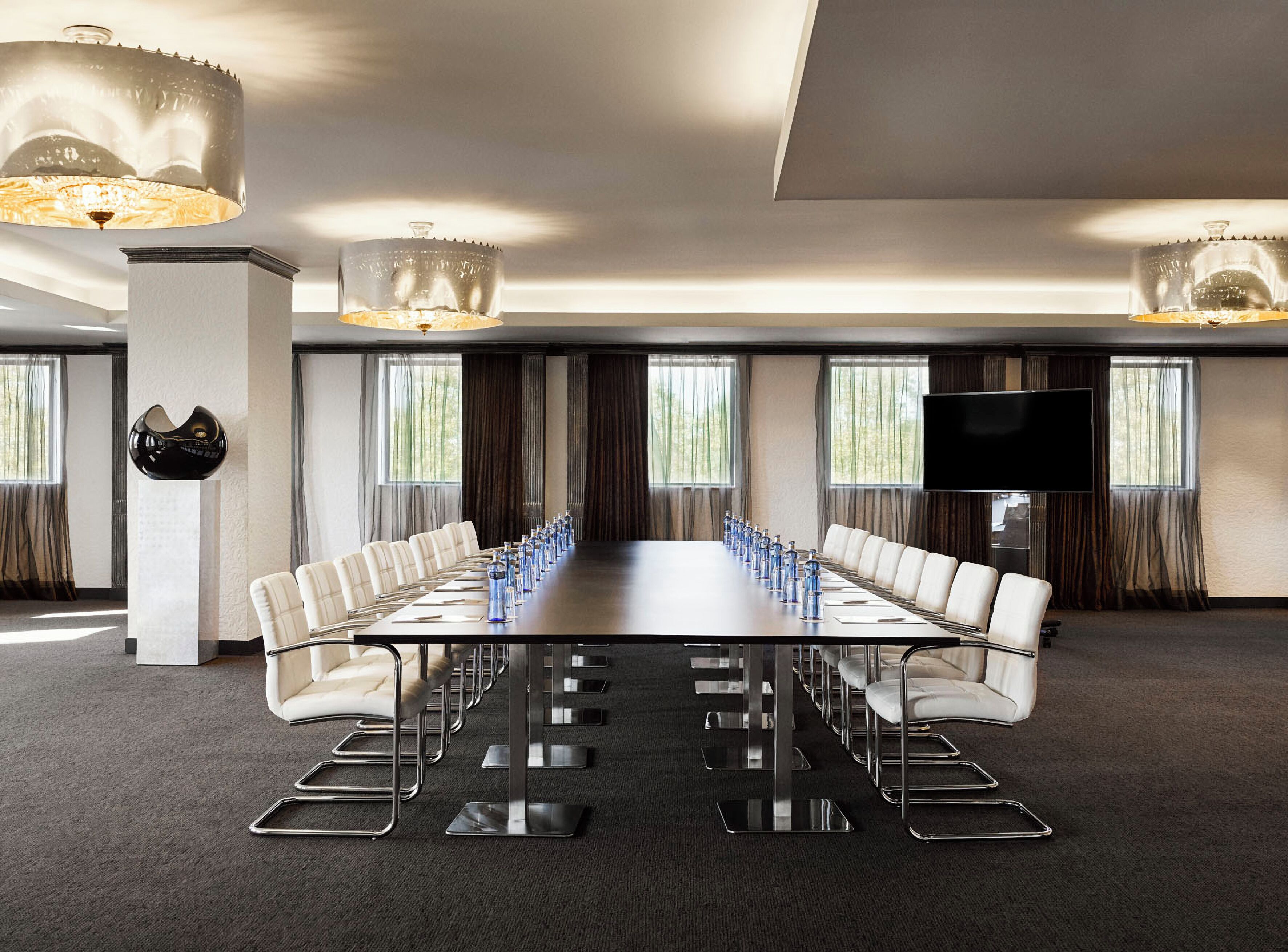 Uma sala de conferências com uma mesa comprida, cadeiras brancas e uma tela de televisão.