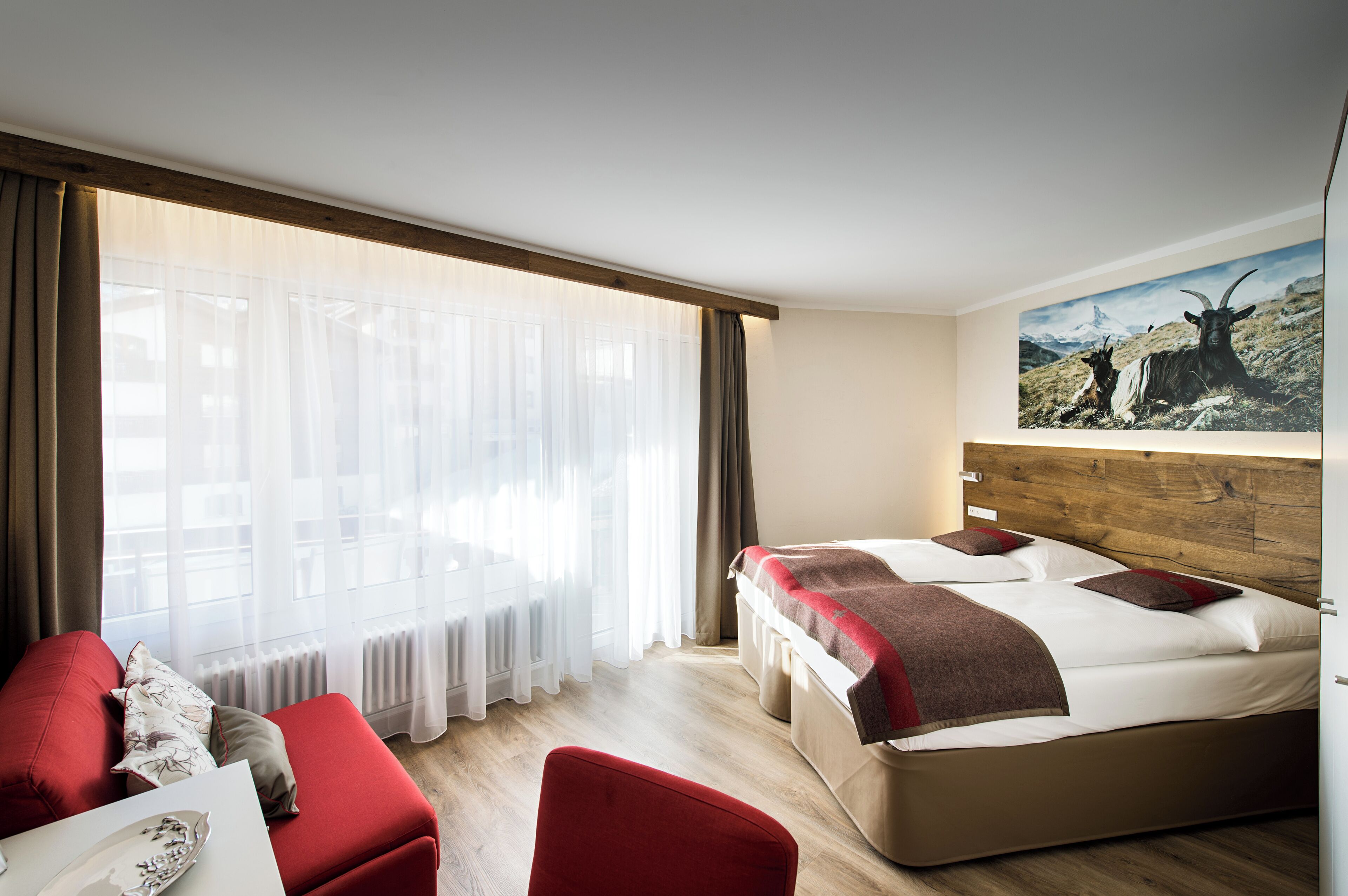 Quarto de hotel com uma cama, um sofá vermelho e um quadro de cabras na parede.