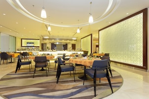 Uma sala de jantar de hotel com mesas redondas, cadeiras e uma cozinha central.