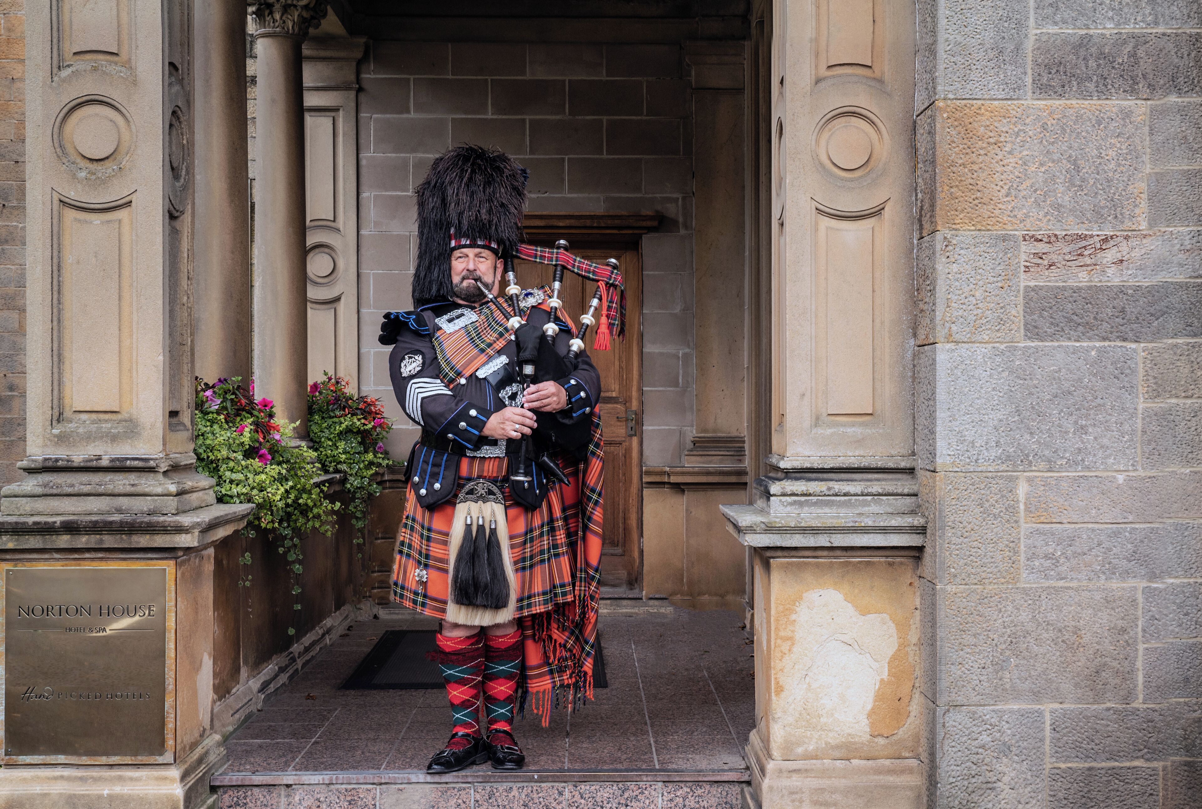 Uma pessoa vestida com traje tradicional escocês em frente à entrada de um edifício com uma placa que diz 'Norton House'.