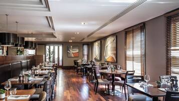 Um restaurante com chão de madeira, várias mesas e cadeiras, e janelas grandes com persianas.