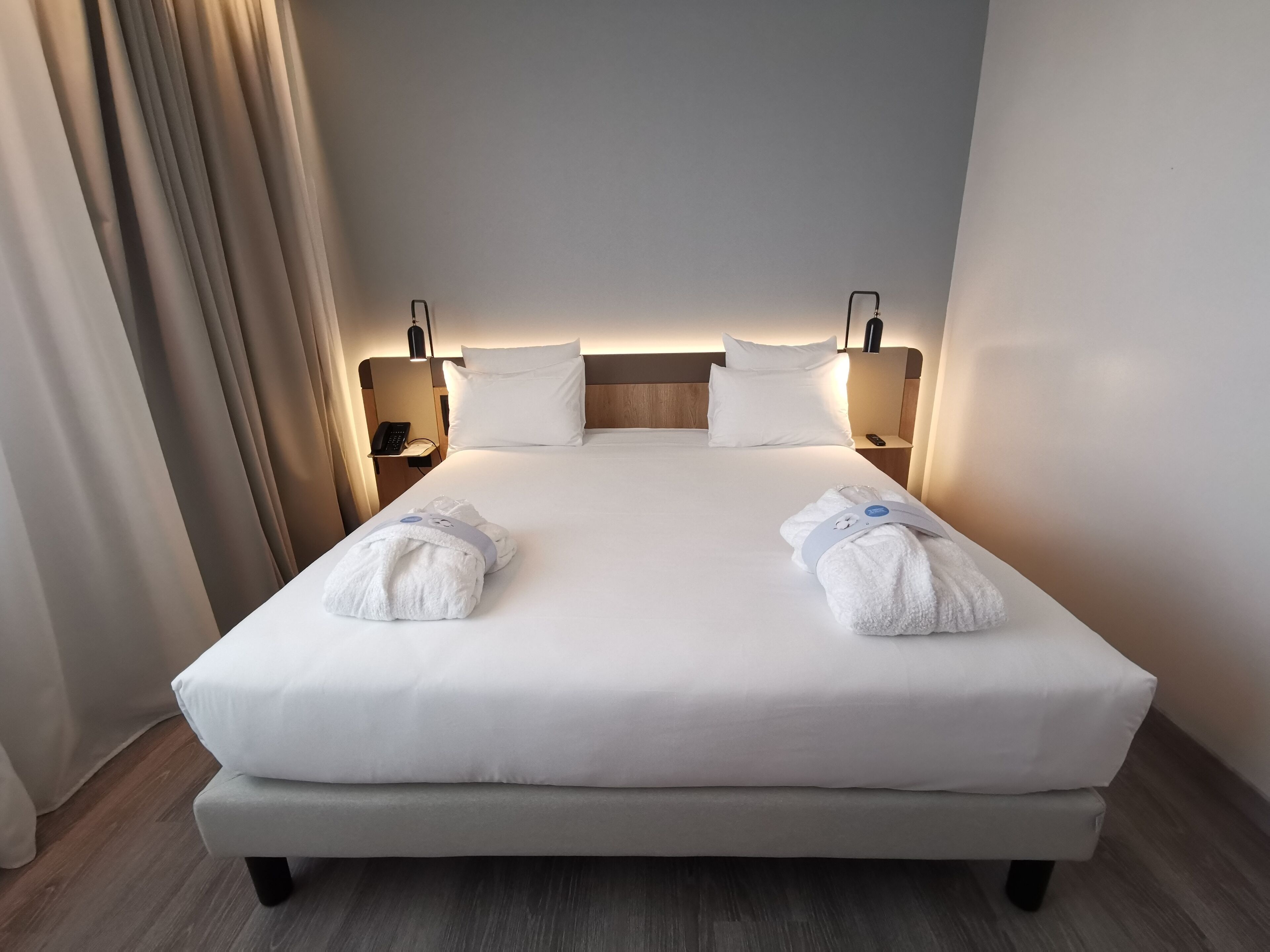 Cama bem arrumada com lençóis brancos e duas toalhas em cada lado. Duas mesas de cabeceira com luminárias e um telefone em cada. O quarto tem piso de madeira e uma cortina bege no lado esquerdo.