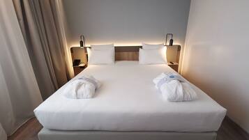 Cama bem arrumada com lençóis brancos e duas toalhas em cada lado. Duas mesas de cabeceira com luminárias e um telefone em cada. O quarto tem piso de madeira e uma cortina bege no lado esquerdo.