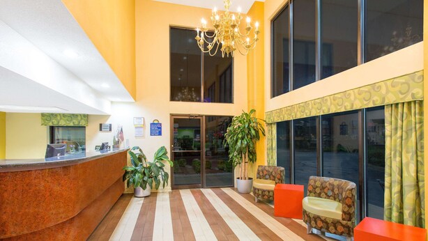 Um saguão de hotel com balcão de recepção, plantas em vasos, área de estar e janelas amplas.