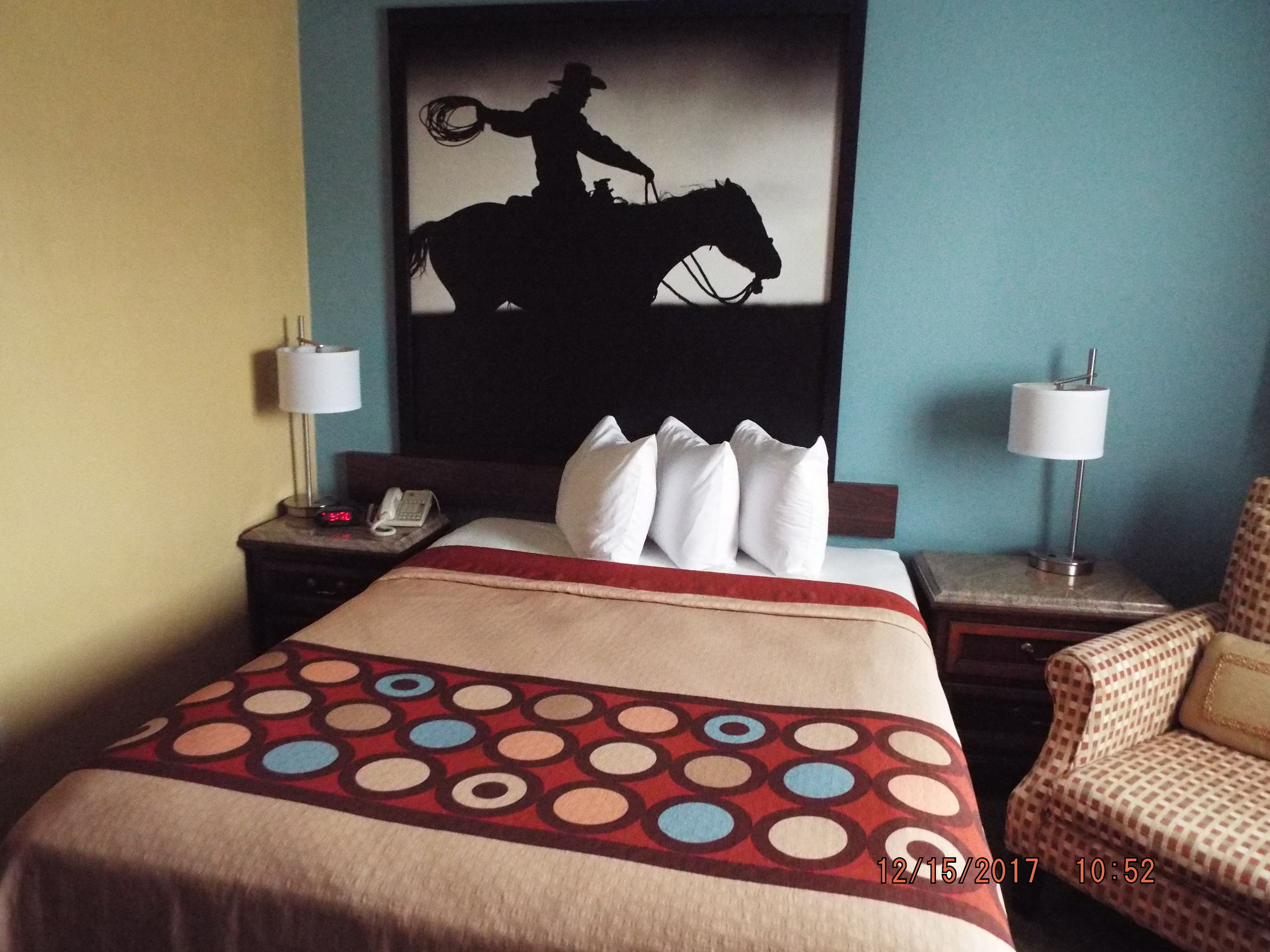 Quarto de hotel com cama, duas mesas de cabeceira, abajur, cadeira e um quadro de um cowboy a cavalo.