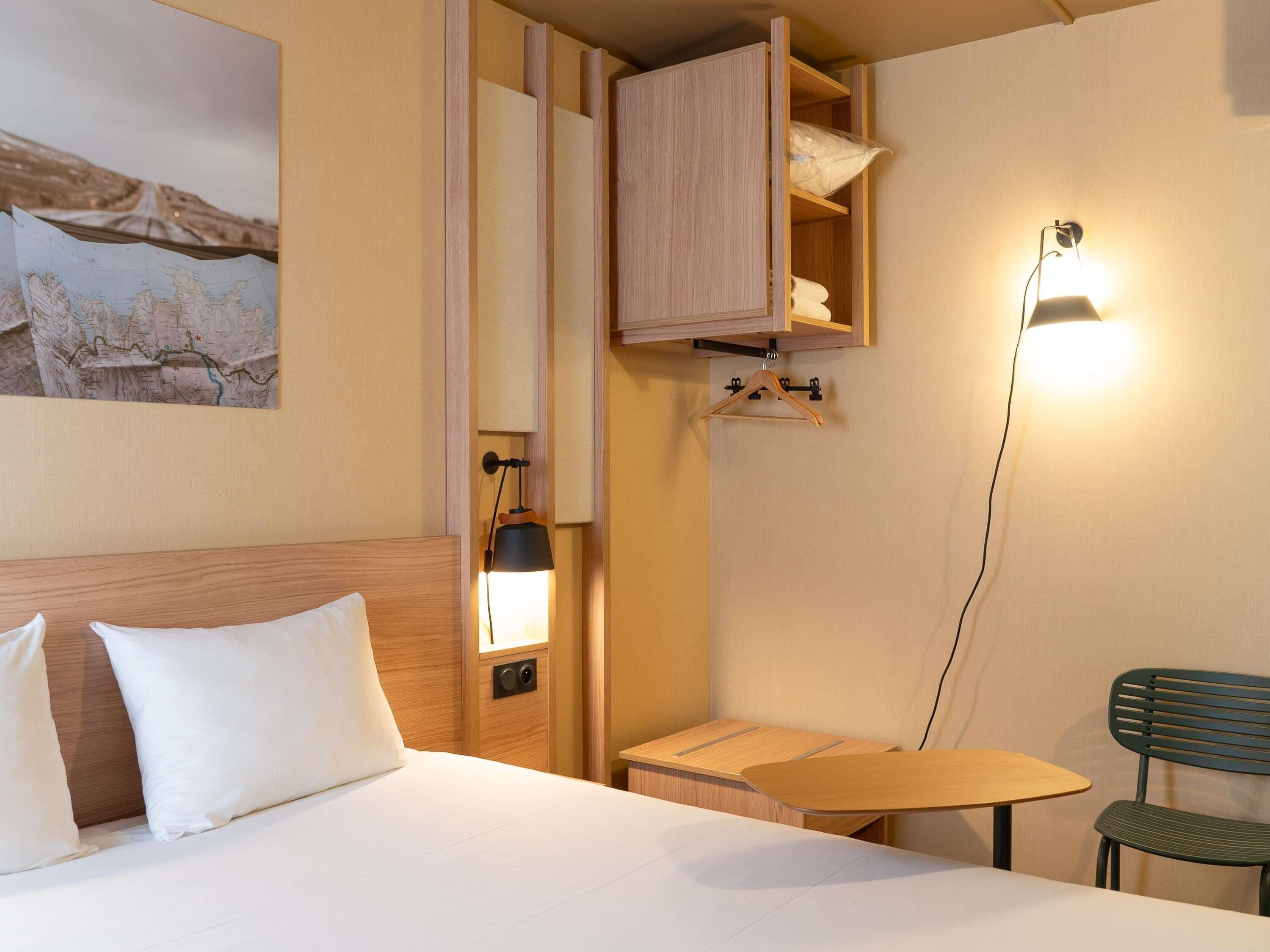 Quarto de hotel com cama, uma pequena mesa, uma cadeira, luminária na parede, mapa e guarda-roupa.