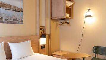 Quarto de hotel com cama, uma pequena mesa, uma cadeira, luminária na parede, mapa e guarda-roupa.