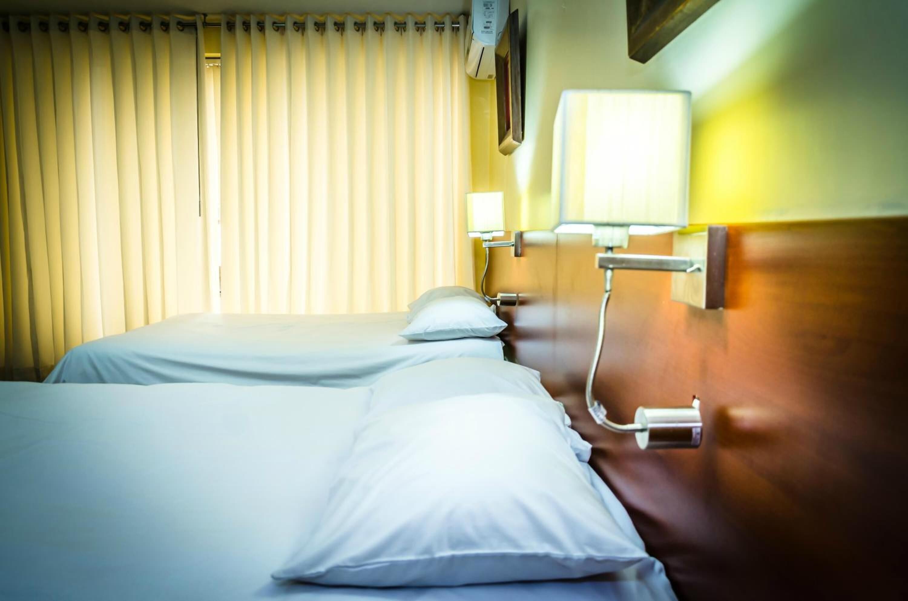 Quarto de hotel com cama, mesa de cabeceira, abajur e janela com persianas.