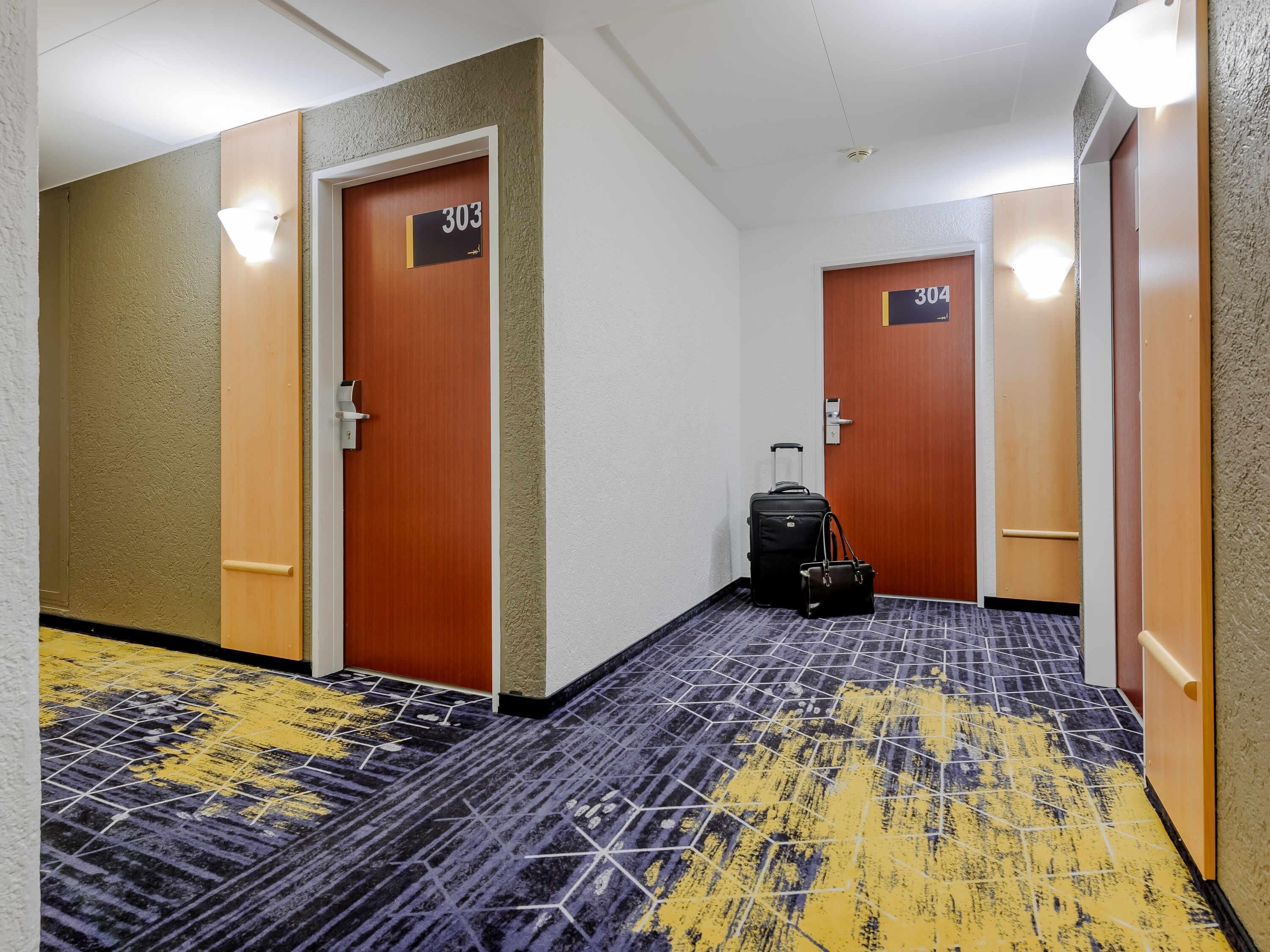 Um corredor de hotel com portas numeradas 303 e 304, um tapete estampado e uma mala preta.