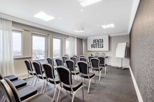 Sala de conferências com fileiras de cadeiras, um quadro branco e uma grande janela com vista para a cidade.