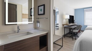 Quarto de hotel com banheiro, destacando um espelho grande, pia e bancada. Há uma escrivaninha com cadeira e televisão.