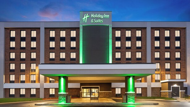 Edifício Holiday Inn & Suites com design moderno, apresentando uma entrada destacada com toldo iluminado em verde e uma placa visível sobre a entrada.