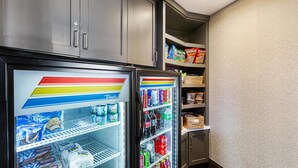 Uma despensa de hotel com um frigorífico cheio de snacks e bebidas, e prateleiras com produtos embalados.