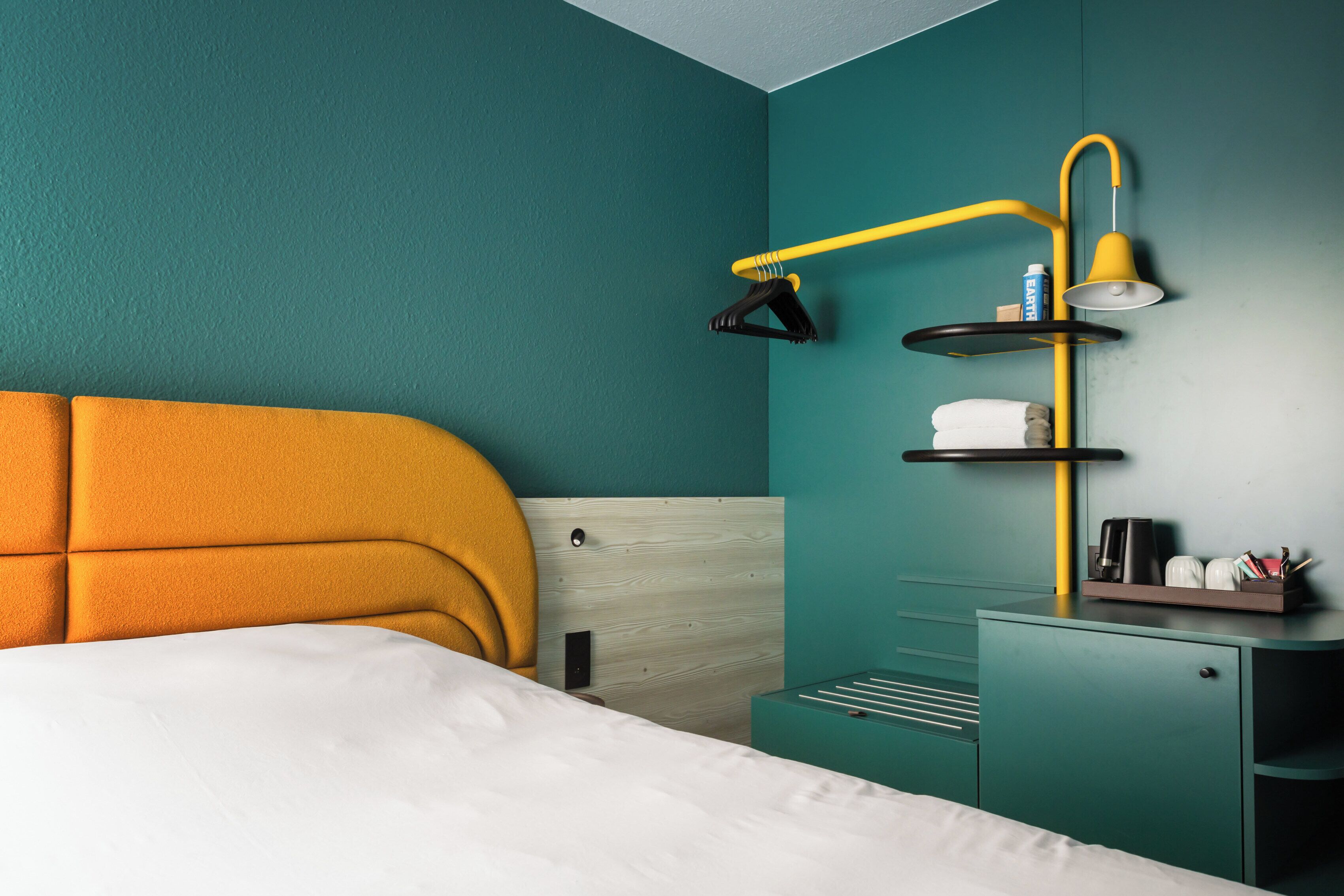 Quarto de hotel moderno com parede em tom de verde-água, cabeceira de madeira, cama com roupa de cama branca, abajur amarelo, prateleira com artigos de higiene pessoal e mesa de cabeceira verde.