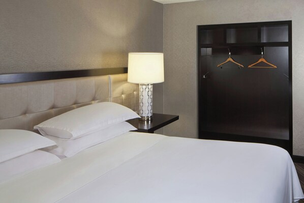 Quarto de hotel com cama, almofadas, mesa de cabeceira com abajur e guarda-roupa com cabides.