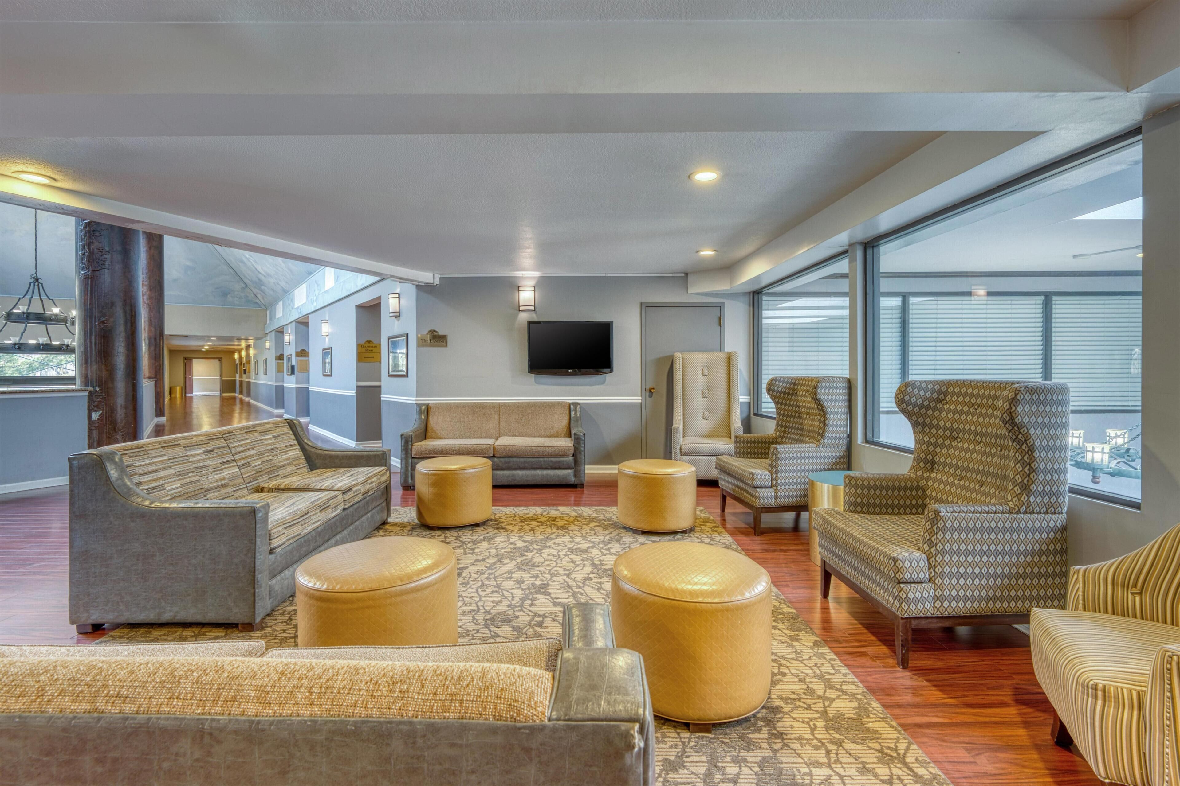 Quarto de hotel moderno com sofá, poltronas, pufes, TV de tela plana e janelas amplas.