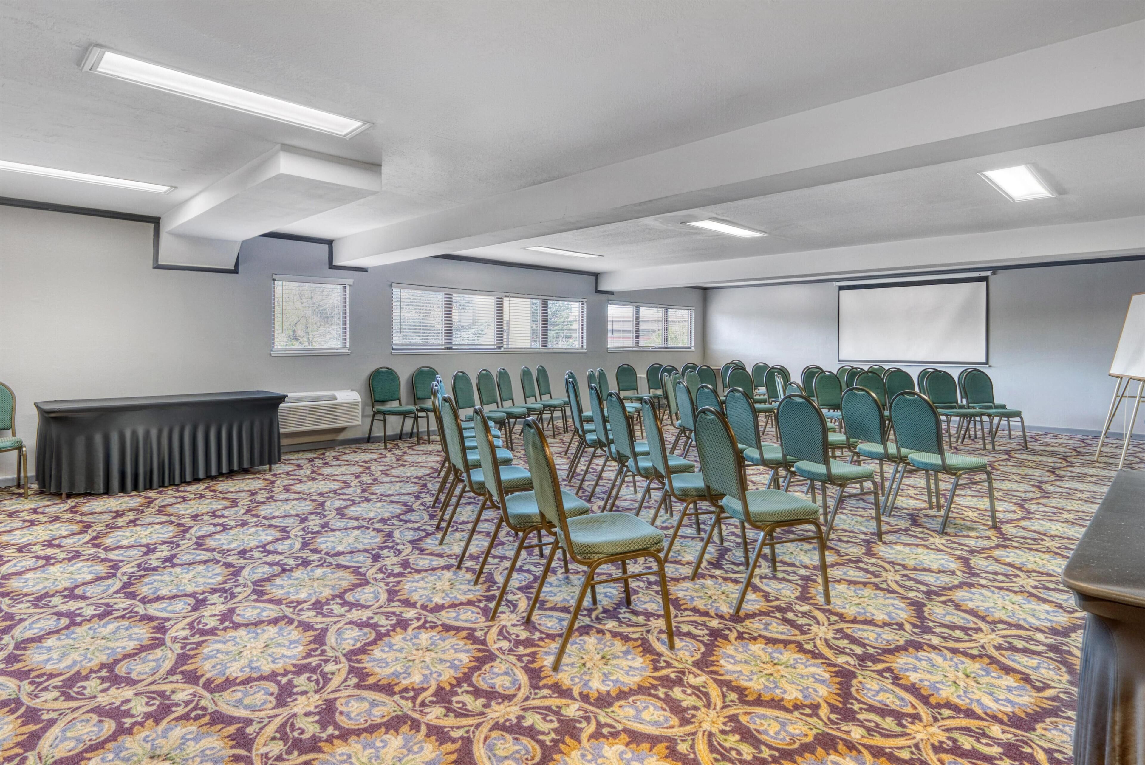Uma sala de conferências com fileiras de cadeiras verdes, piso coberto de carpete, uma tela de projetor e uma mesa com toalha preta.