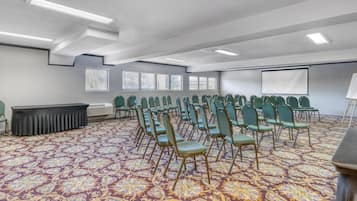 Uma sala de conferências com fileiras de cadeiras verdes, piso coberto de carpete, uma tela de projetor e uma mesa com toalha preta.