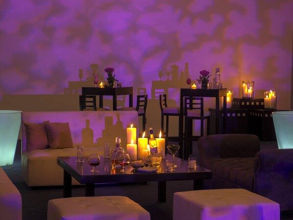 Uma área de estar pouco iluminada, com sofá, mesa de centro e mesa de jantar posta com velas e copos.