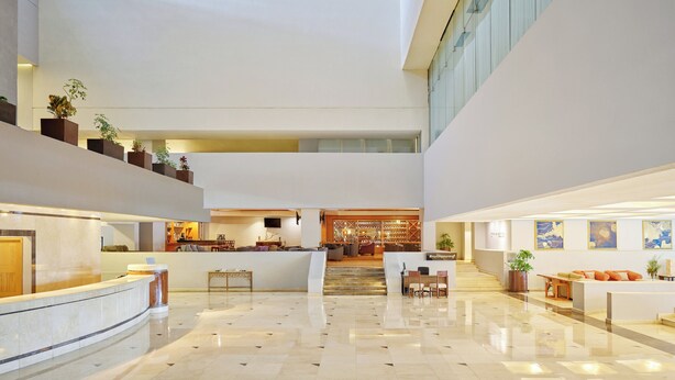 Um hall amplo com teto alto, balcão de recepção, áreas de estar e escada que dá acesso aos andares superiores.