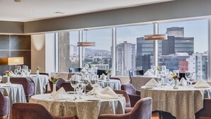 Restaurante com mesas redondas preparadas para refeições, amplas janelas com vista para a cidade e iluminação pendente moderna.