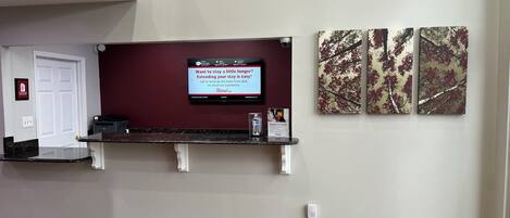 Uma área de recepção de hotel com uma tela digital exibindo uma mensagem sobre prolongar a estadia, um balcão de atendimento e três obras de arte emolduradas na parede.