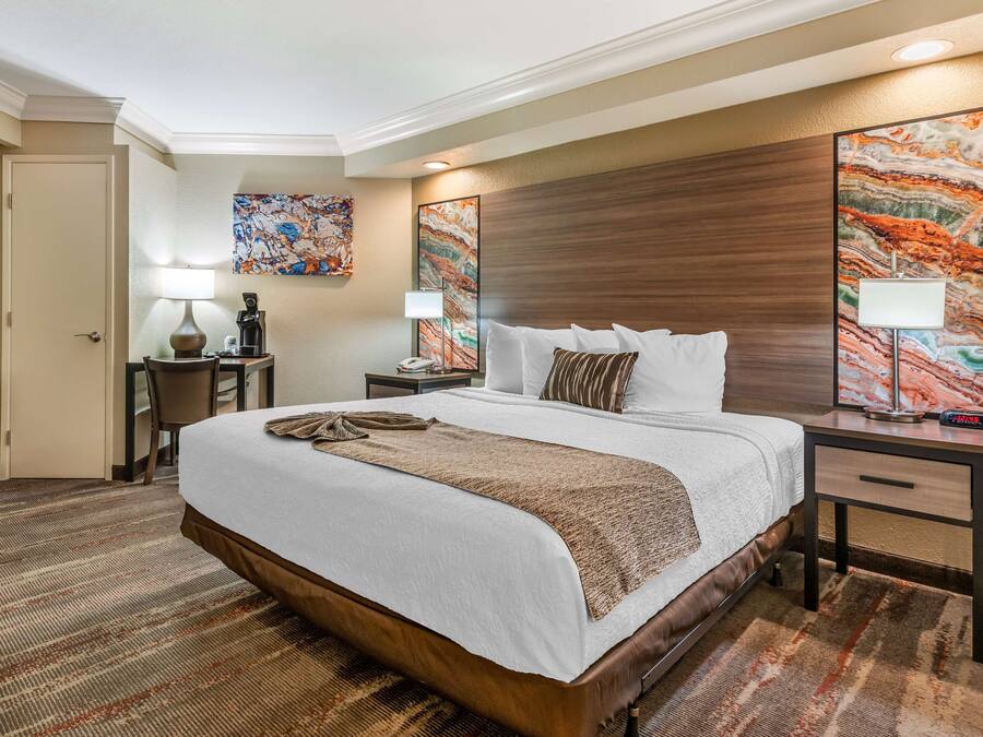 Quarto de hotel com uma cama grande, duas mesas de cabeceira, uma escrivaninha e uma cadeira. Há duas luminárias fixadas na parede e um quadro sobre a cama.