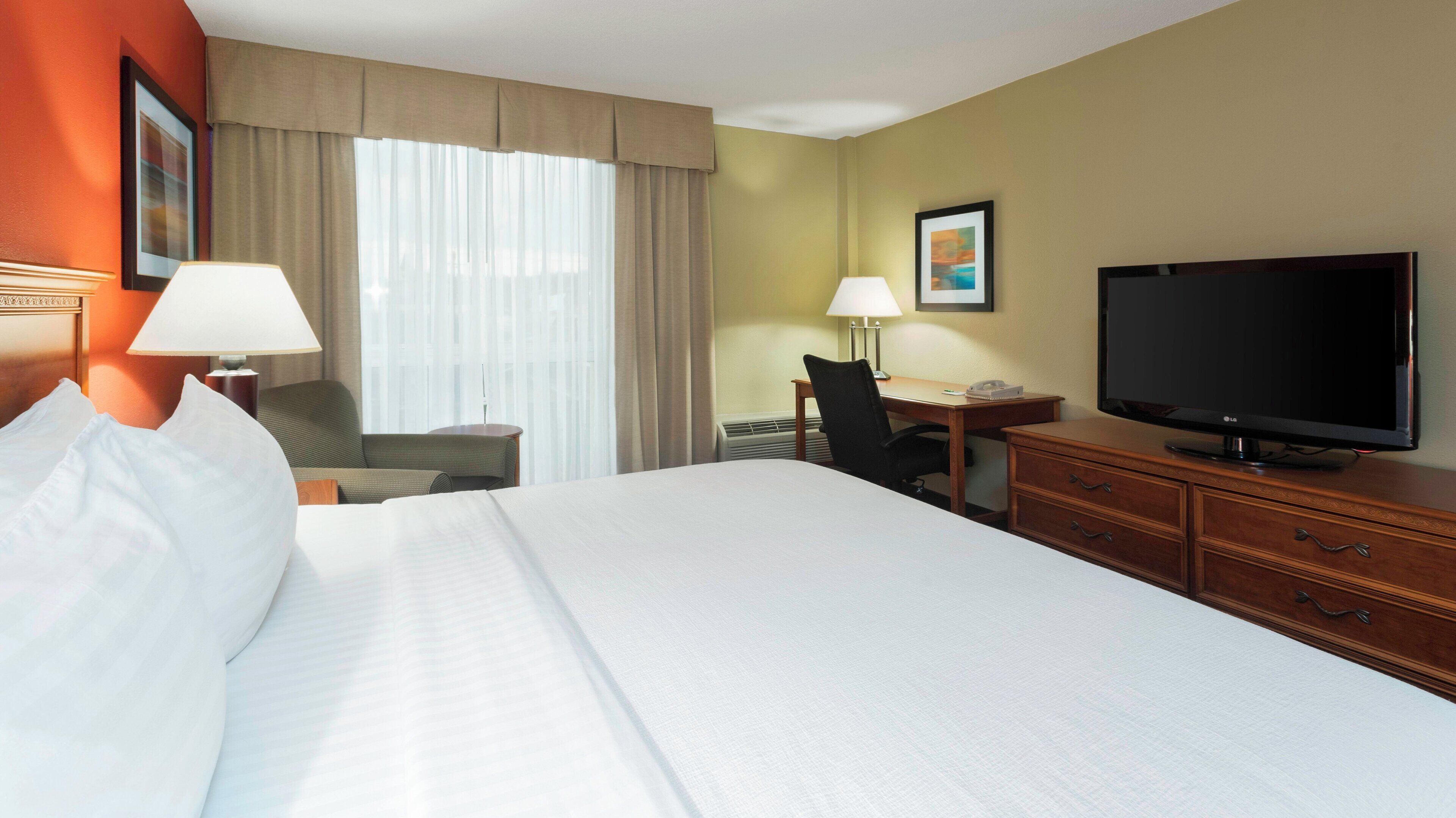 Quarto de hotel com uma cama, uma escrivaninha com cadeira, uma televisão e um abajur.
