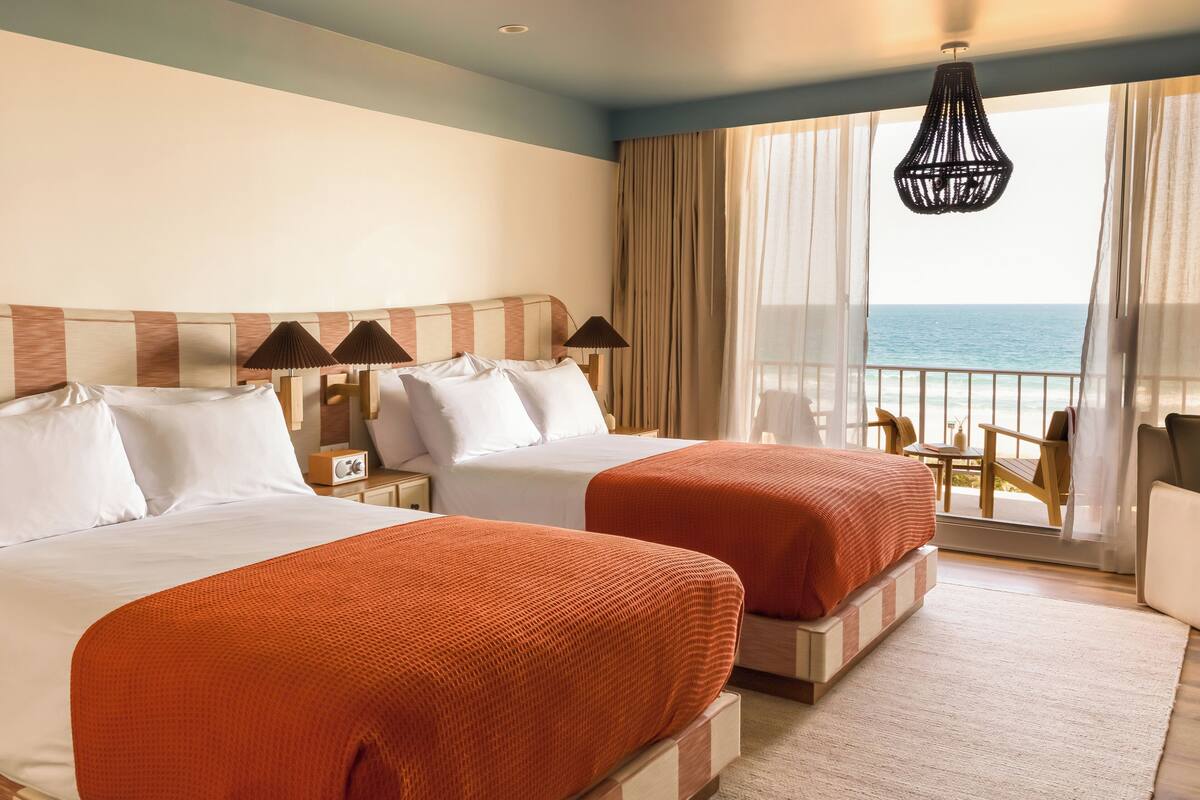 Quarto de hotel com duas camas, uma janela grande com vista para o mar, uma varanda com cadeiras e um lustre.