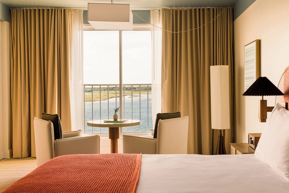 Quarto de hotel com uma cama grande, duas cadeiras, uma mesinha, um abajur e vista para a água pela janela.