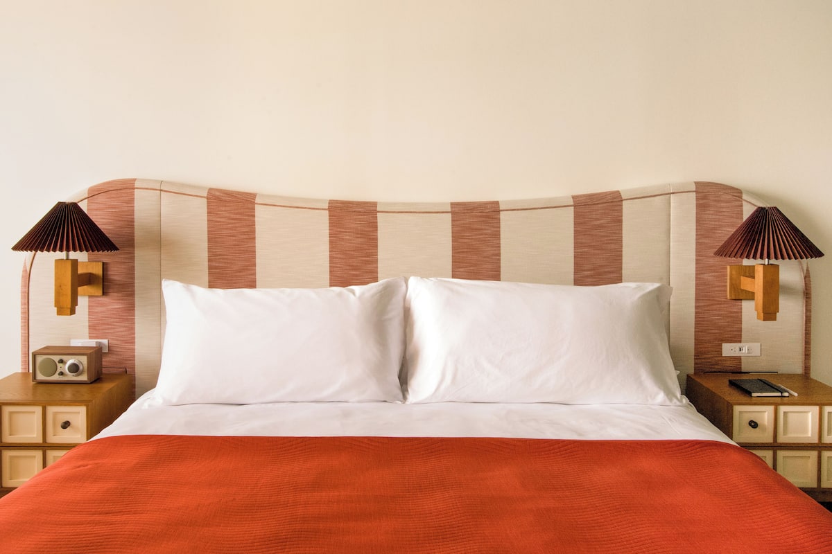 Uma cama bem arrumada com almofadas brancas e um cobertor vermelho, ladeada por duas mesas de cabeceira com abajures e um rádio.