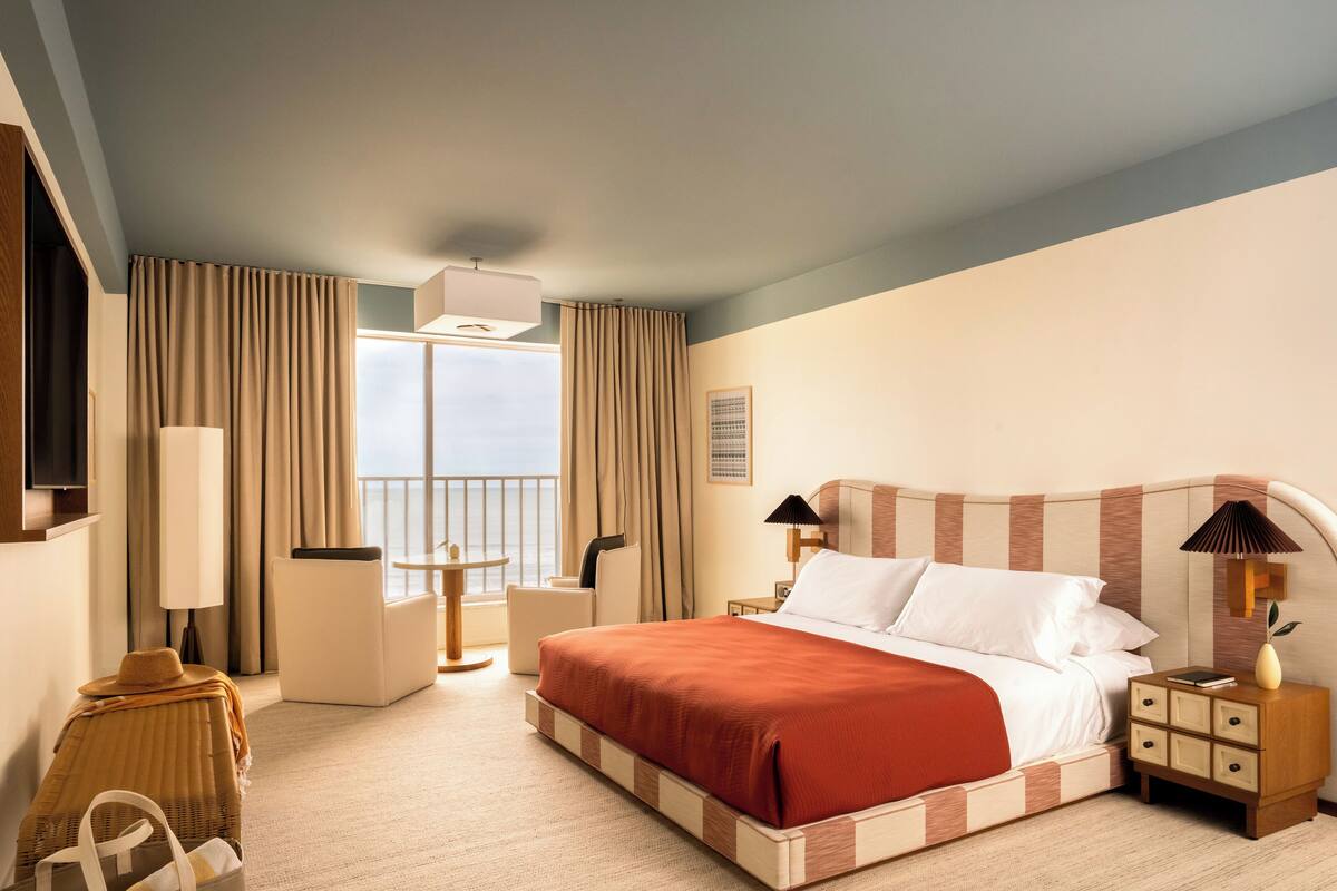 Quarto de hotel com uma cama grande, mesa de cabeceira, cadeira, escrivaninha, abajur e vista para o mar.