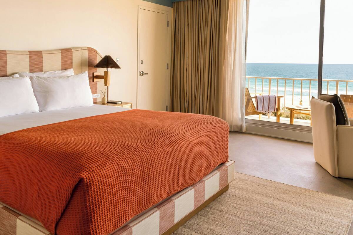 Quarto de hotel com uma cama grande, mesa de cabeceira, luminária, vista para o oceano e varanda com uma cadeira.