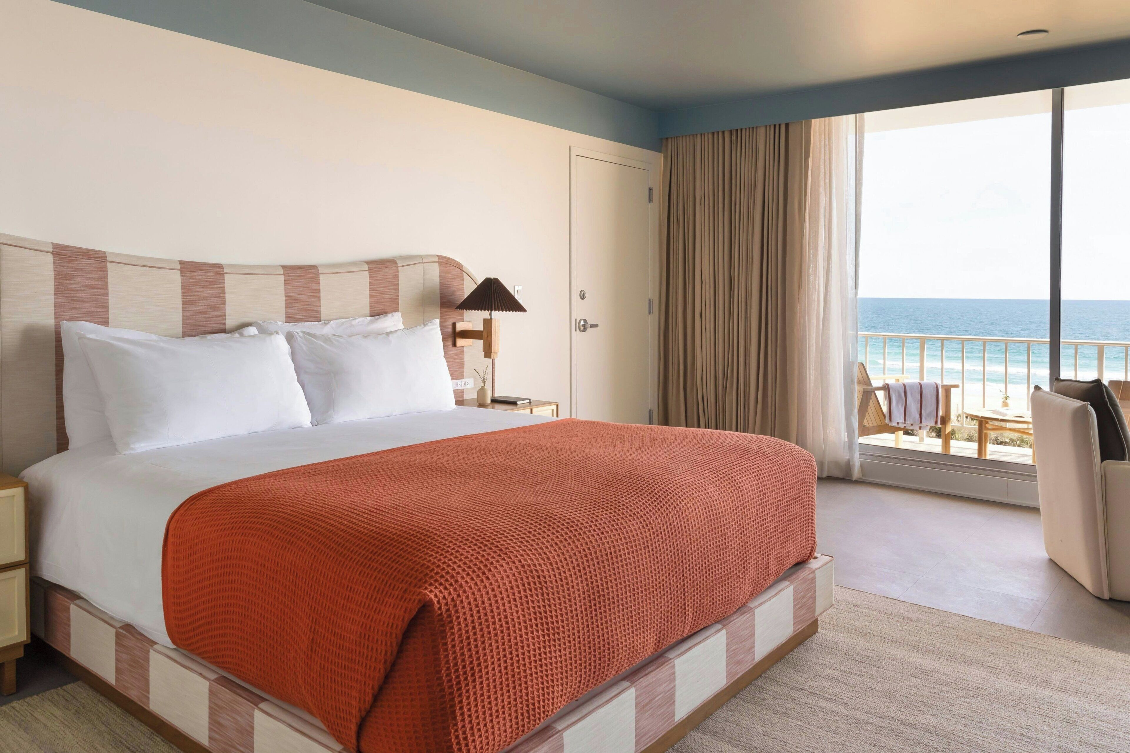Quarto de hotel com uma cama grande, uma mesa de cabeceira, uma luminária, vista para o oceano através de uma varanda e uma porta.