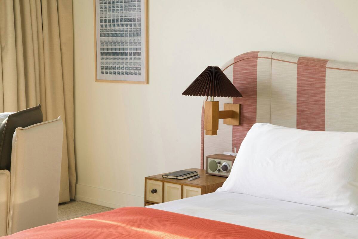 Quarto de hotel com cama, mesa de cabeceira com rádio, cadeira e quadro pendurado na parede.
