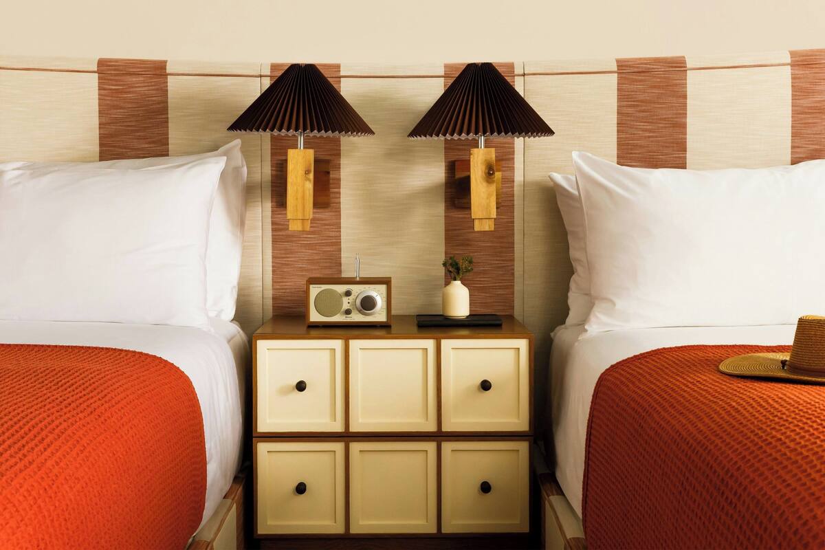 Quarto de hotel com duas camas, cada uma com lençóis brancos e cobertores laranja. Há duas mesas de cabeceira, com luminárias e um rádio.